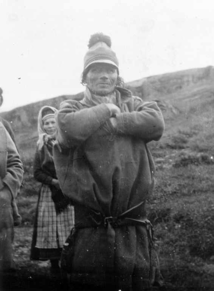 Mann med samedrakt og armene i kors. Kvinne i samedrakt bak. Kofte ...