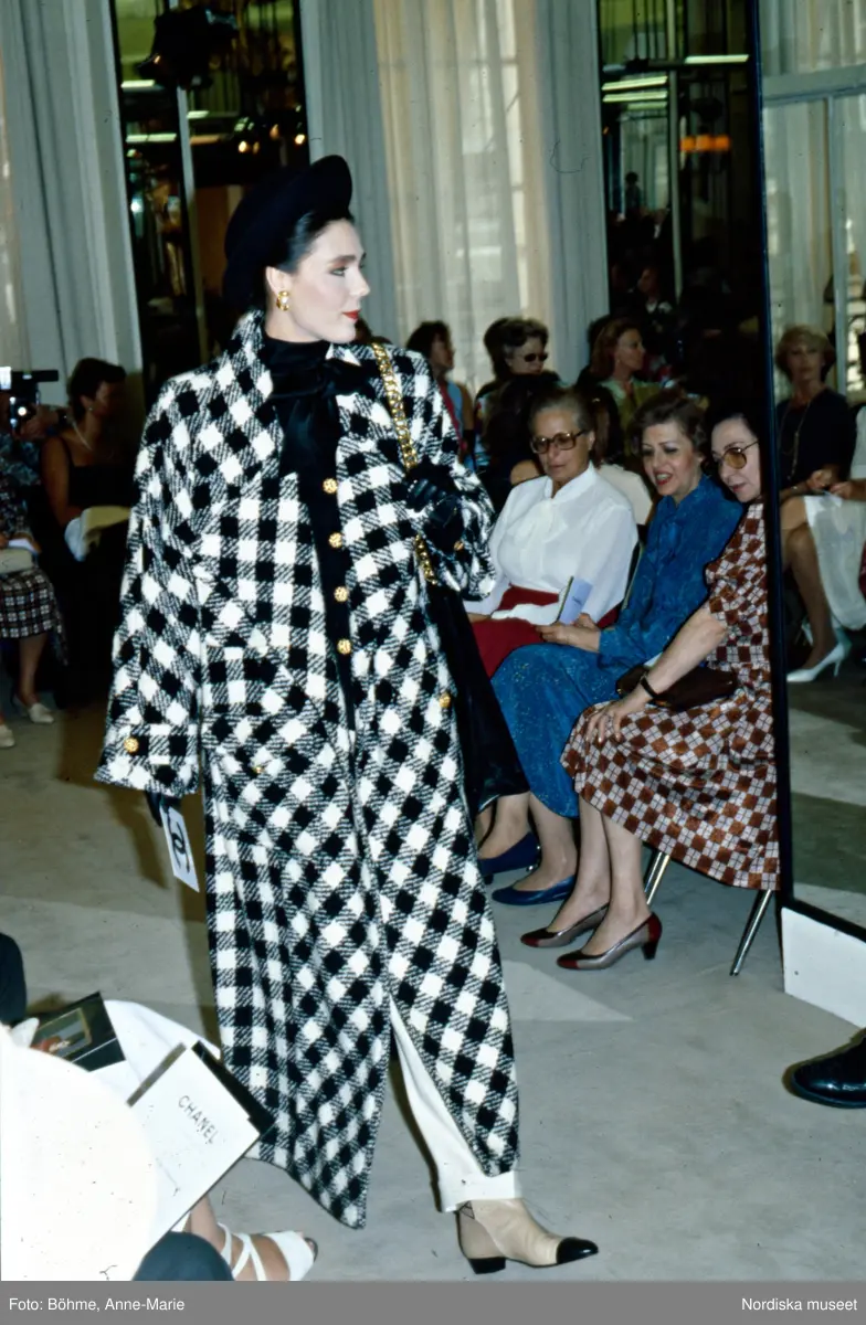 Modevisning. Modell i rutig, lång kappa i svart och vitt, axelväska med ...