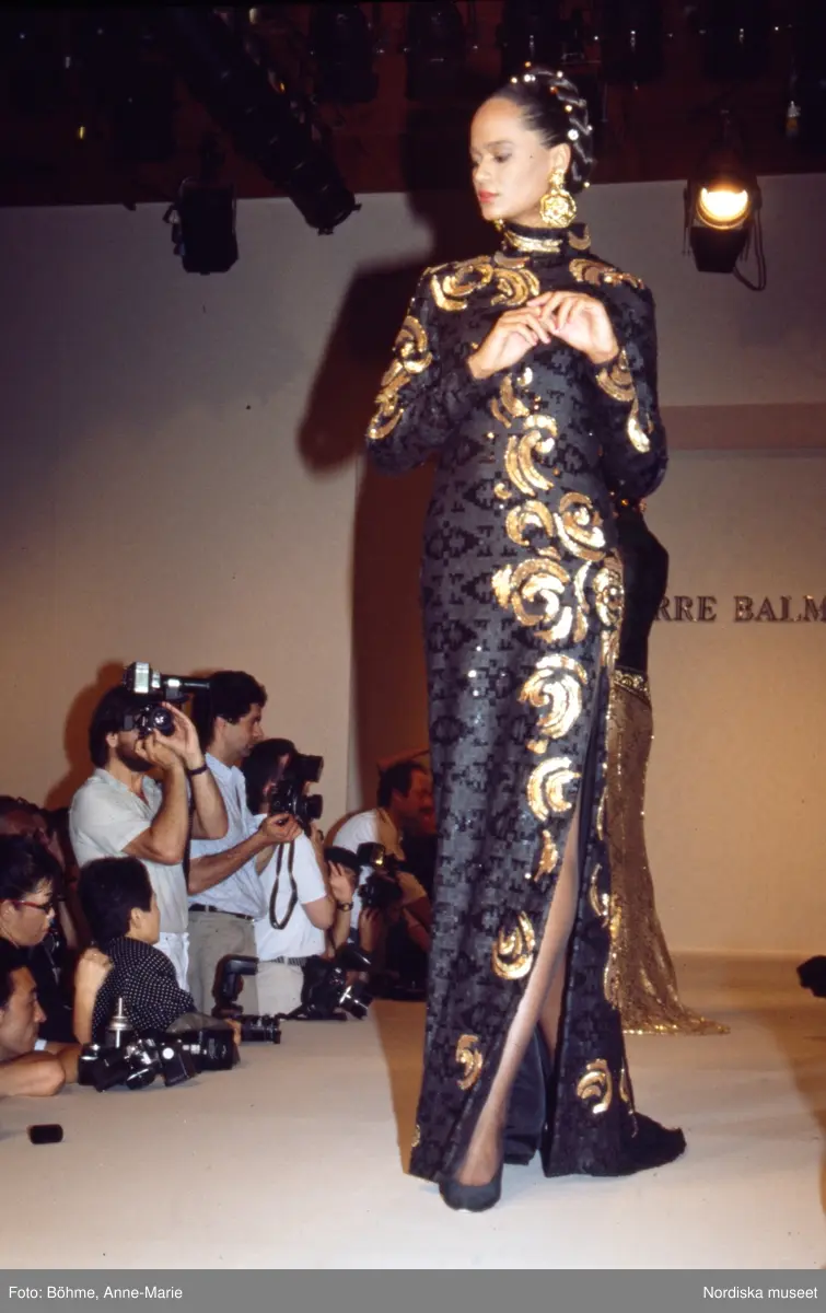 Modevisning. Modell i svart långklänning med slits och ...