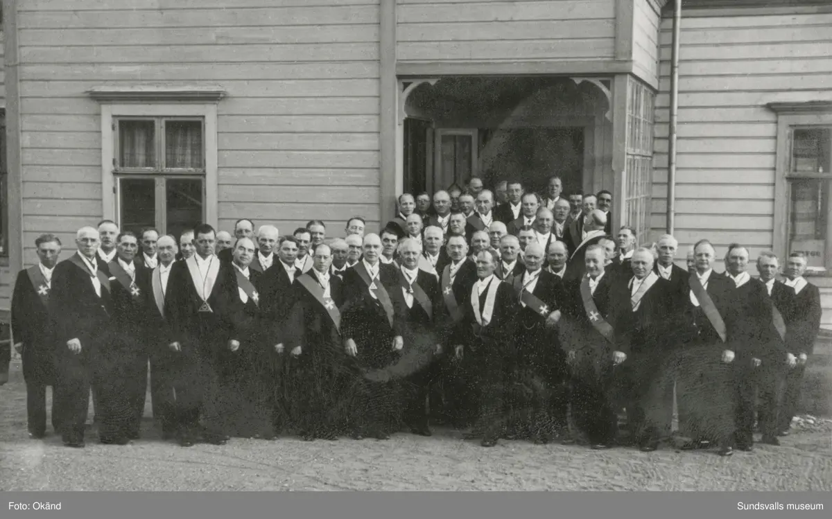 Gruppfoto utanför en större träbyggnad, en stor grupp män, många med ...