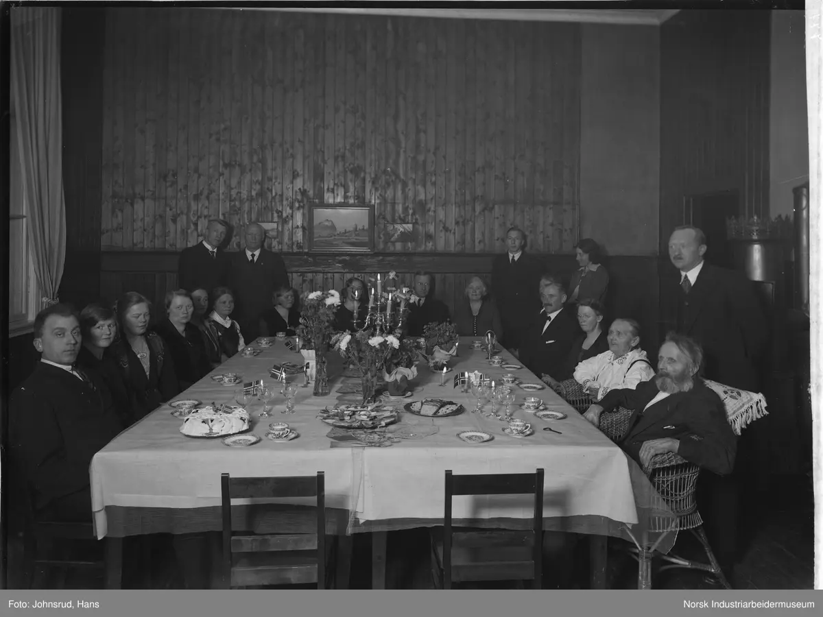 Gruppe rundt dekket bord. Bordet er pyntet med flagg og blomster. Kaker ...