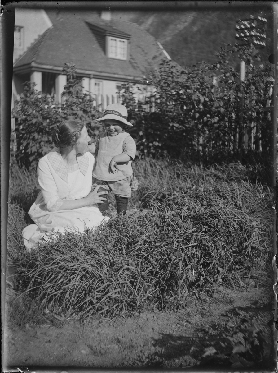 Kvinne sittende i hage i Rødbyen med barn ved siden av seg. Barnet har ...
